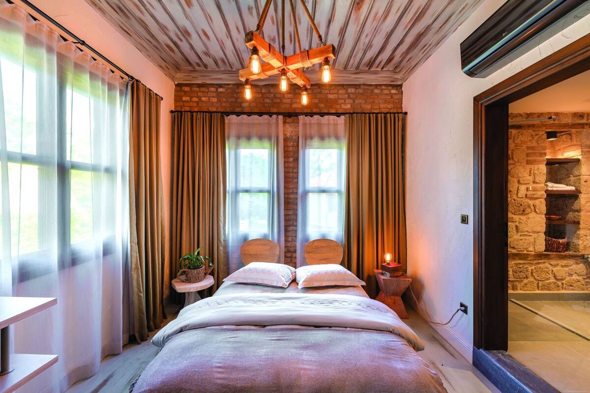 Quarto com uma cama, cabeceira de madeira, mesa de cabeceira com abajur e vista para uma parede de pedra.