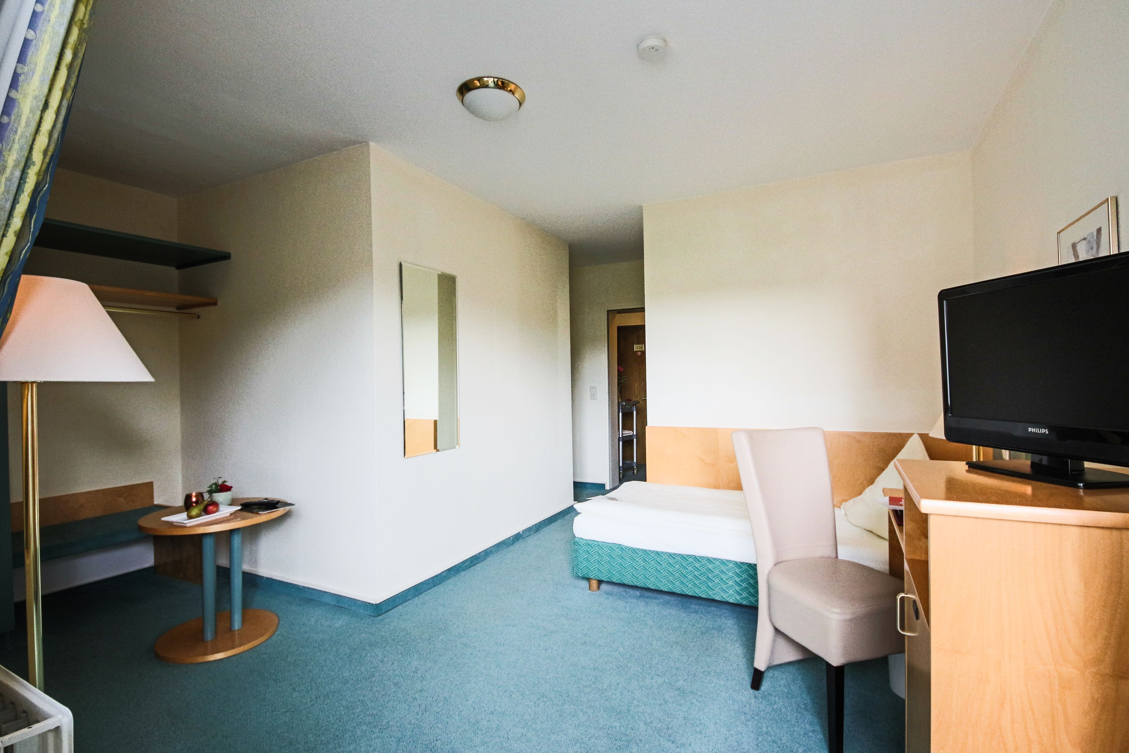 Quarto de hotel com cama, televisão, escrivaninha com luminária, cadeira e mesinha com frutas.