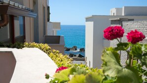 Varanda costeira com vista para o mar, apresentando um edifício branco, um muro de pedra e uma fileira de flores cor-de-rosa.