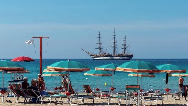 Uma praia com fileiras de guarda-sóis e espreguiçadeiras, um navio grande ao fundo e céu azul claro.