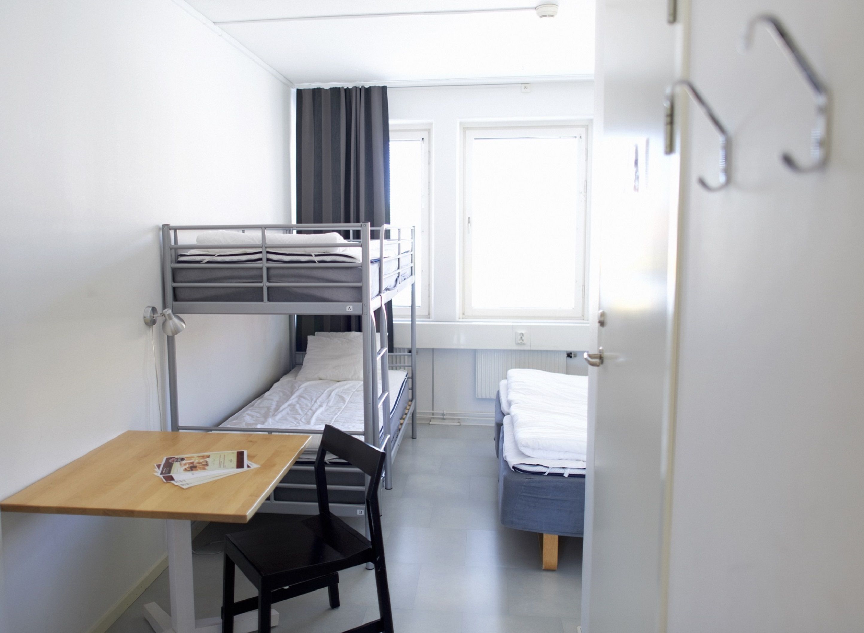 Quarto em dormitório com beliches, uma mesinha, uma cadeira e uma janela com cortinas.