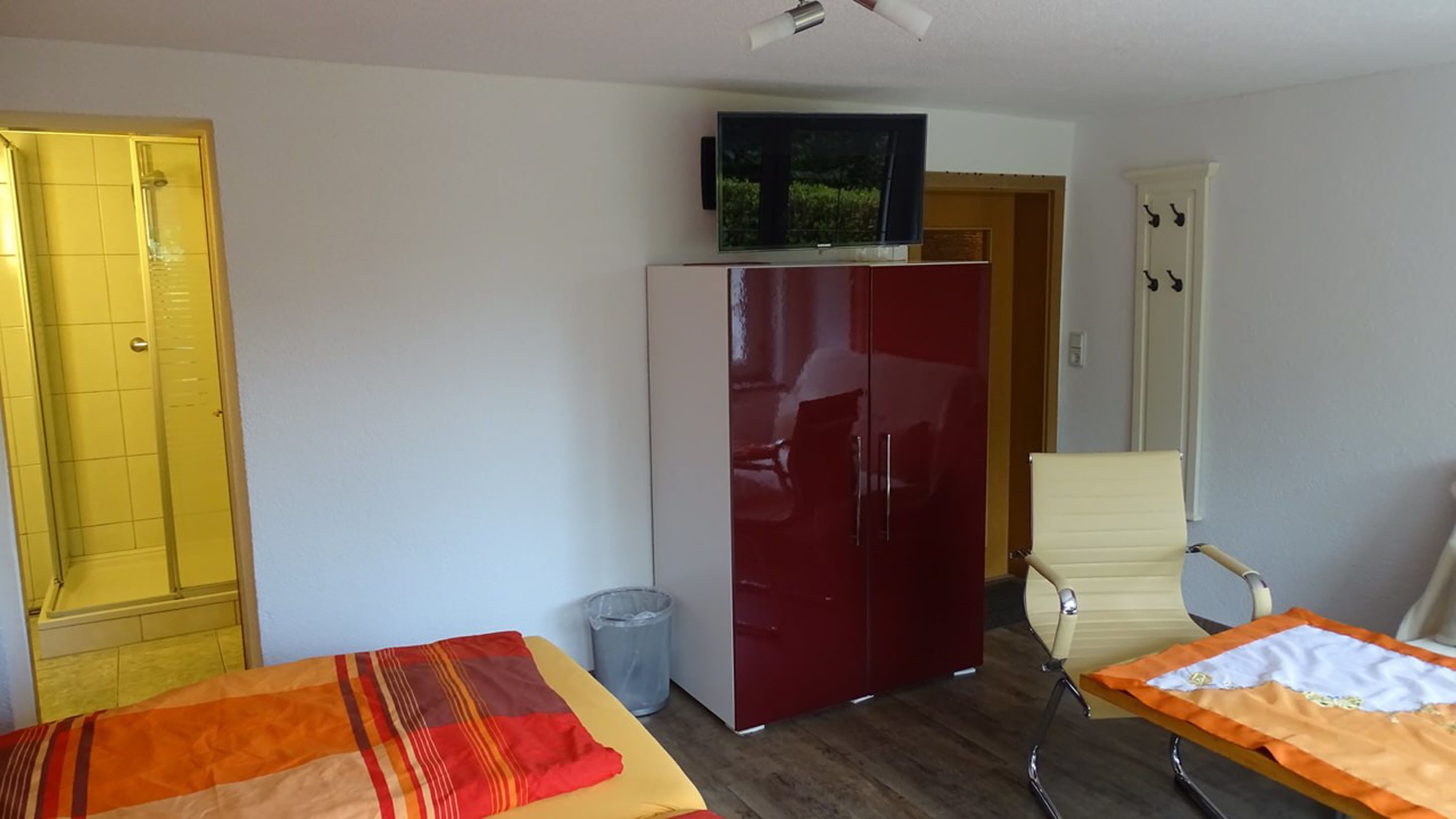 Quarto com cama, guarda-roupa vermelho, televisão por cima, uma cadeira e uma porta.