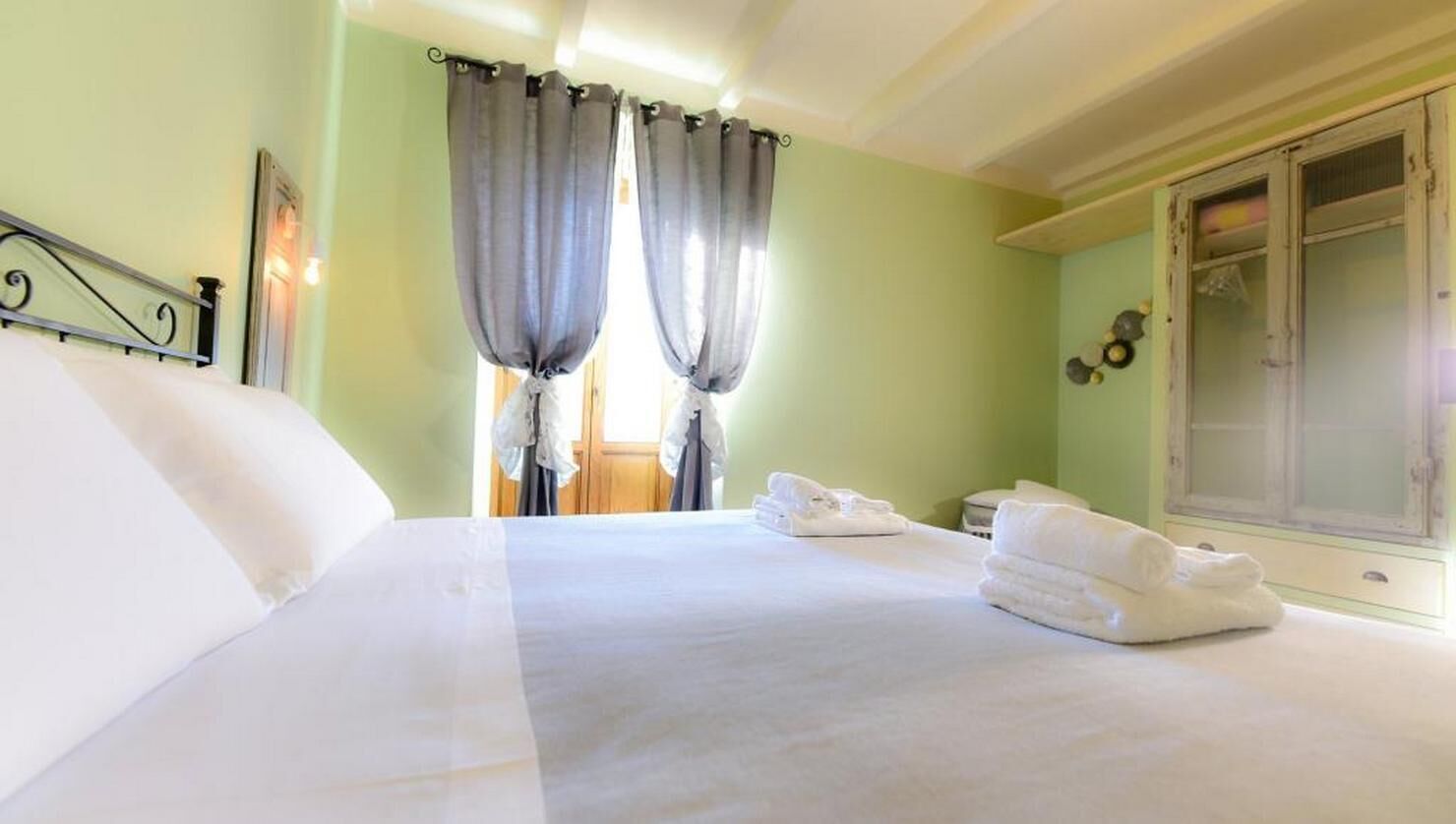 Uma cama bem arrumada com lençóis brancos, uma janela com cortinas e uma mesa de cabeceira com toalhas.
