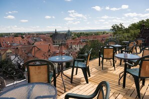 Terraço no telhado com área de estar externa, oferecendo vista de uma cidade histórica com edifícios de telhado vermelho e o topo de uma igreja.