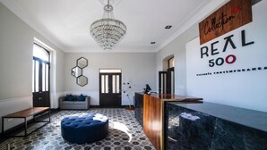 Um hall de hotel moderno com balcão de recepção, um lustre, um banquinho azul e um chão com padrão geométrico.