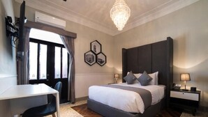 Quarto com uma cama grande, uma escrivaninha, uma cadeira, mesa de cabeceira com abajur e um candelabro.