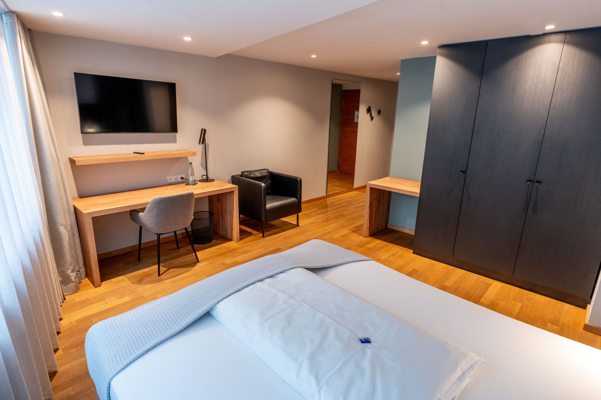 Quarto de hotel moderno com uma cama grande, uma escrivaninha com cadeira, um sofá pequeno, uma televisão e um guarda-roupa.