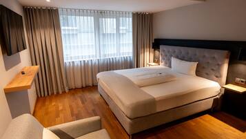 Quarto de hotel moderno com uma cama grande, mesa de cabeceira de madeira, TV na parede e uma janela com cortinas.