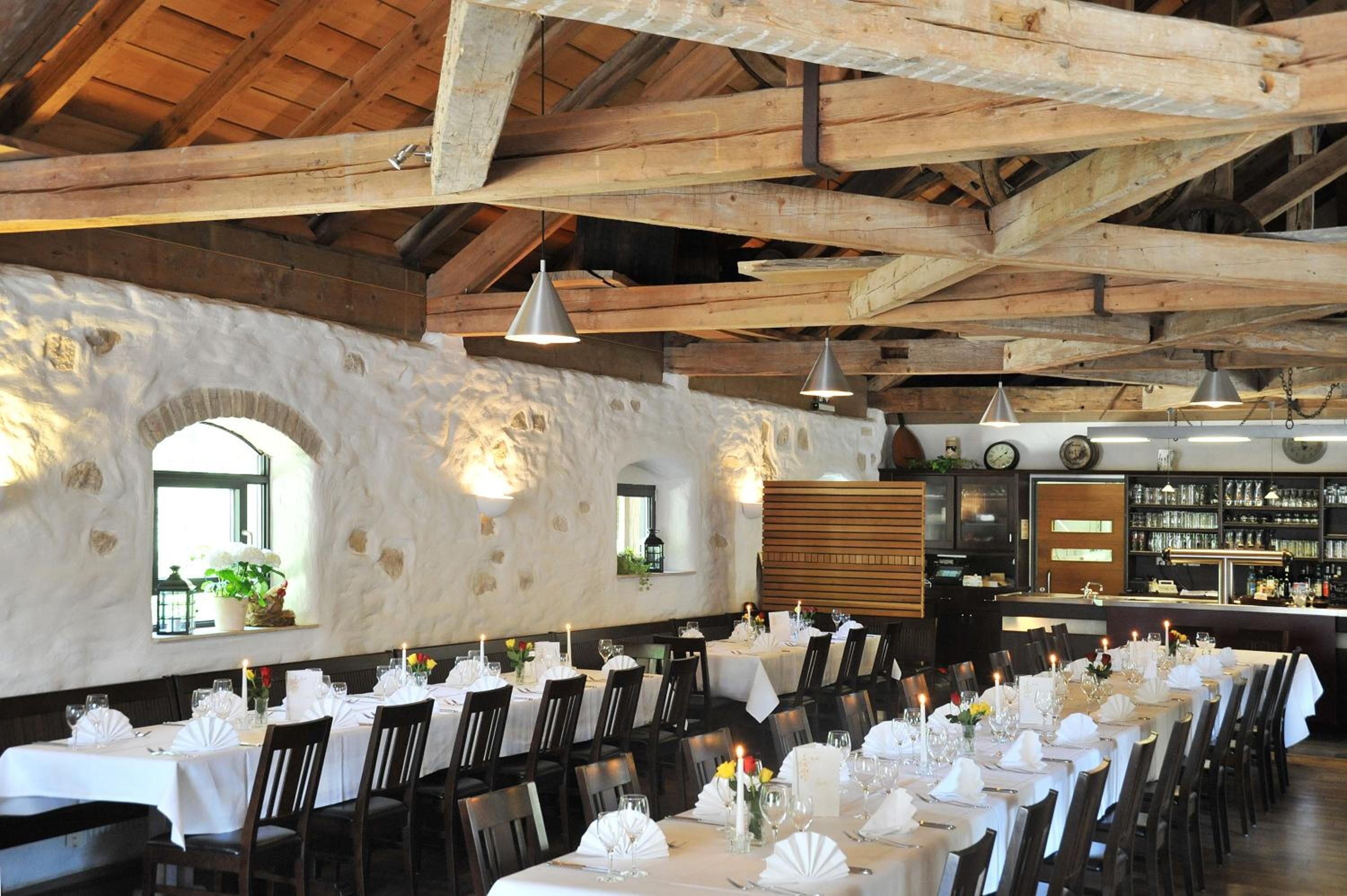 Restaurante com mesas compridas preparadas para refeições, vigas de madeira no teto e área de bar com prateleiras cheias de garrafas.