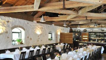 Restaurante com mesas compridas preparadas para refeições, vigas de madeira no teto e área de bar com prateleiras cheias de garrafas.