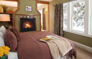 Quarto com lareira, cama com cobertor vermelho, janela com cortinas e vista para árvores lá fora.