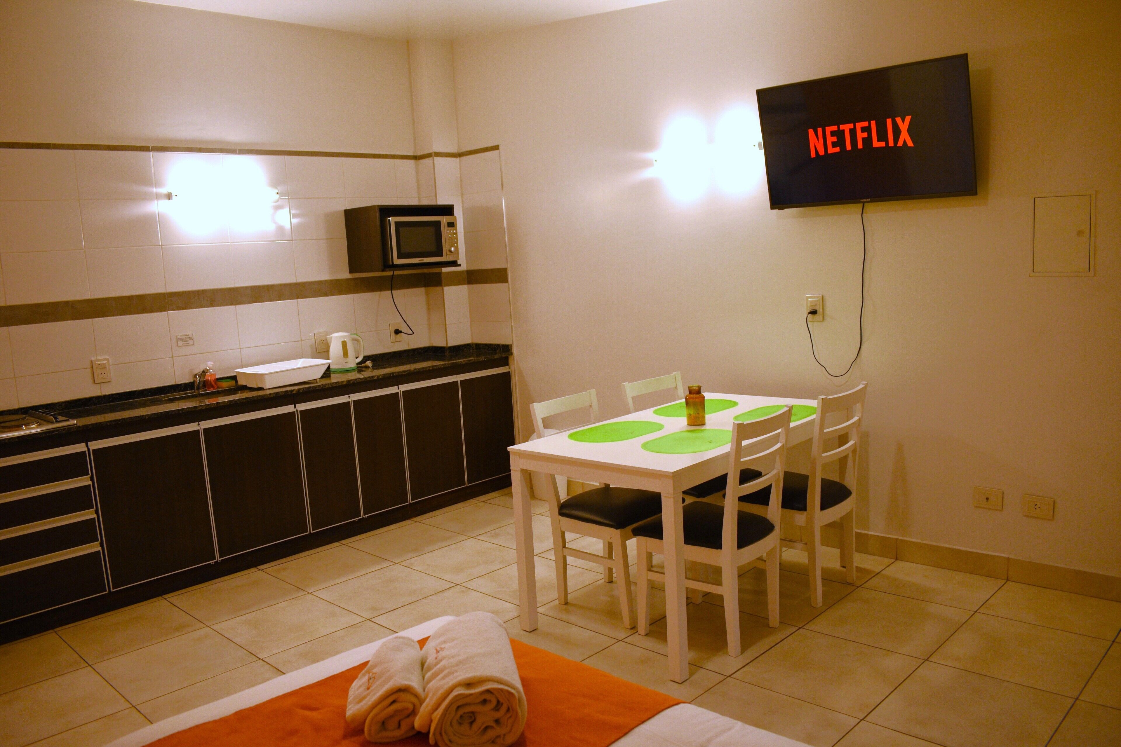 Uma cozinha equipada com micro-ondas, uma mesa de jantar com cadeiras e uma TV na parede exibindo "NETFLIX".