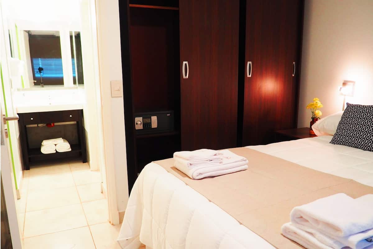 Quarto de hotel com cama, guarda-roupa, banheiro com espelho e mesa de cabeceira com abajur e flores.