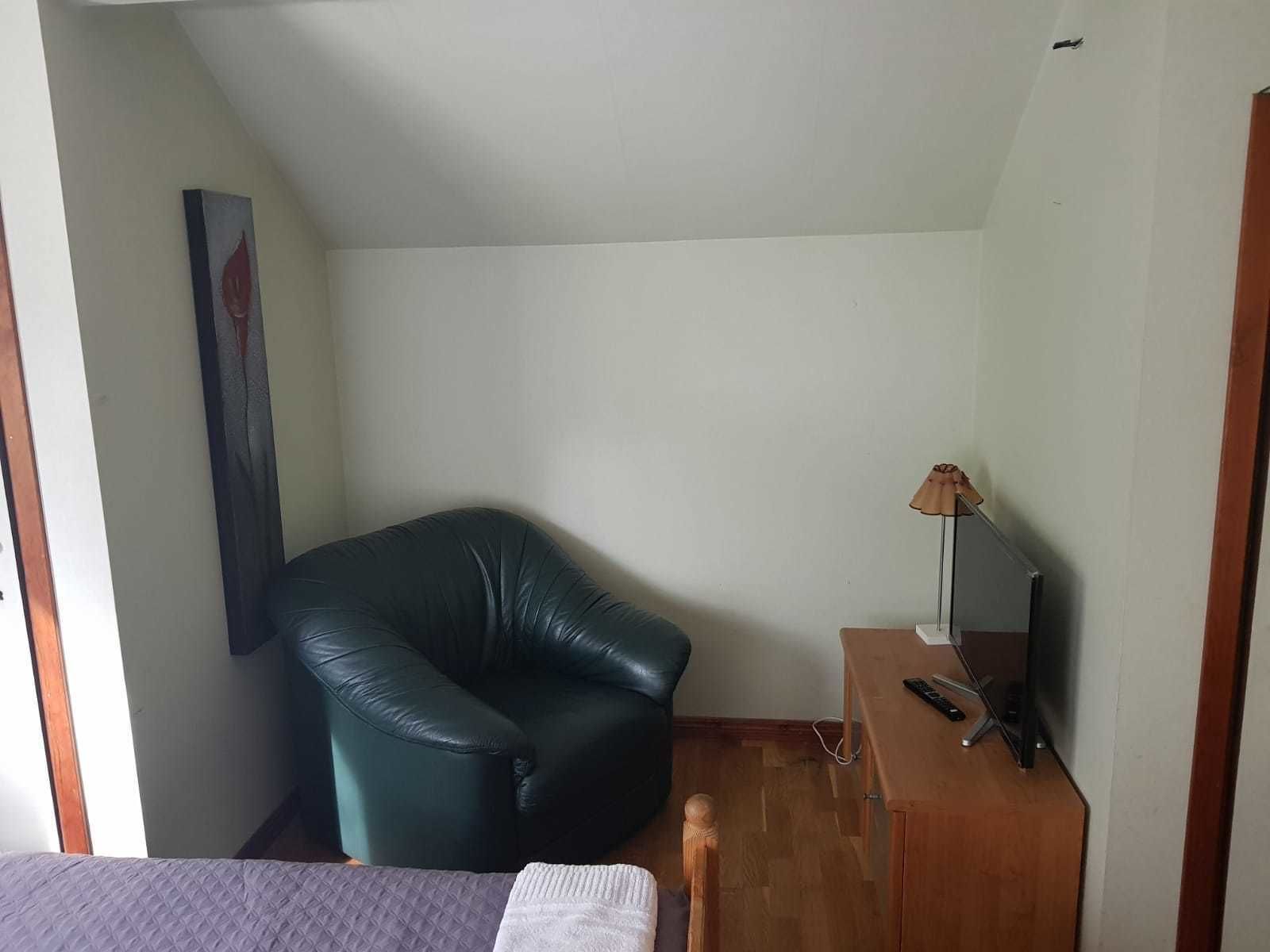 Um quarto pequeno com uma única cadeira de couro preta, uma mesinha de cabeceira de madeira com uma luminária e uma televisão.