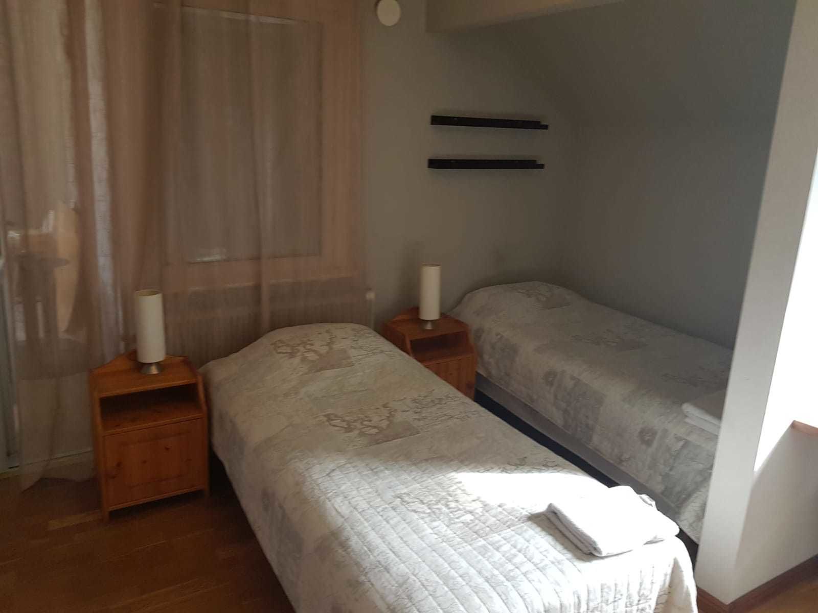 Quarto com duas camas, uma mesinha de cabeceira de madeira com abajur e uma janela com cortinas leves.