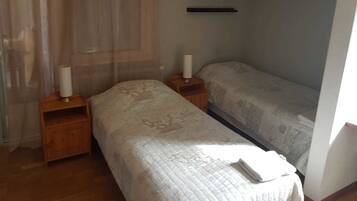 Quarto com duas camas, uma mesinha de cabeceira de madeira com abajur e uma janela com cortinas leves.