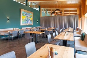 Uma sala de jantar moderna com várias mesas e cadeiras, um quadro grande na parede e vista para o exterior através de grandes janelas.