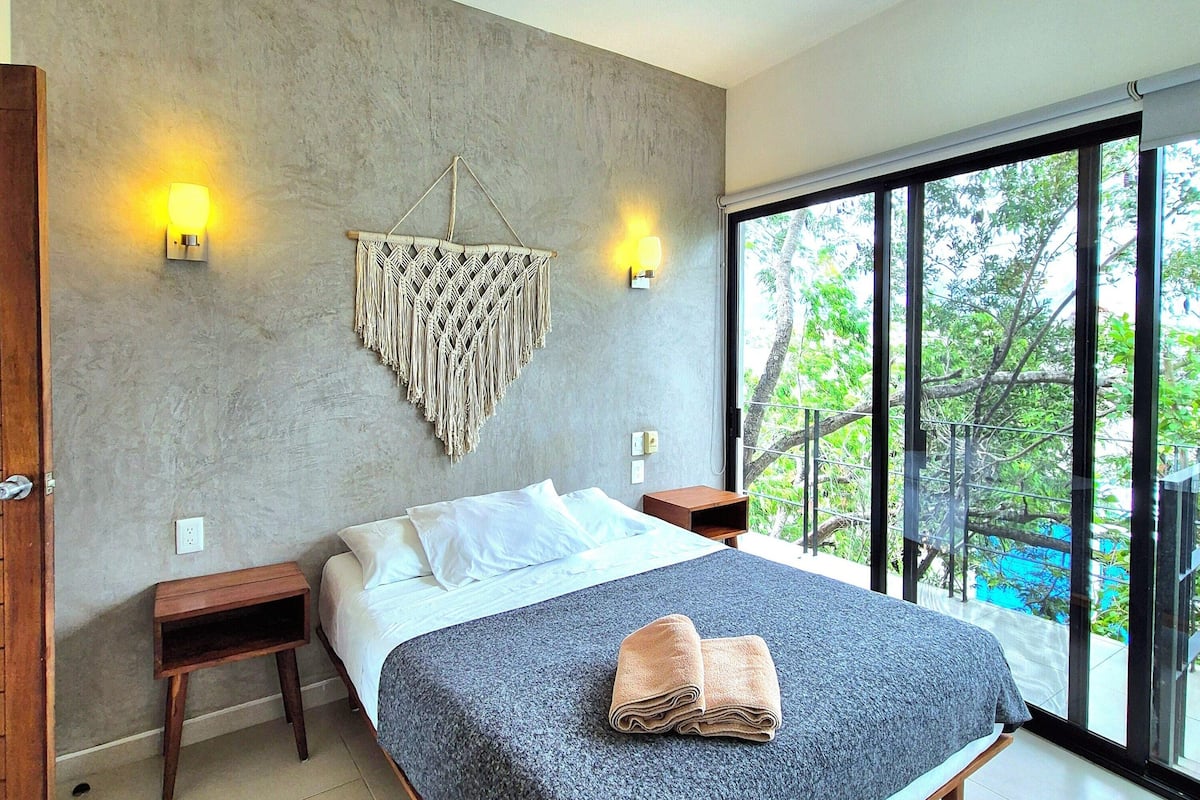 Quarto moderno com uma cama grande, duas mesas de cabeceira, um ventilador de teto e vista para o verde através de grandes janelas.