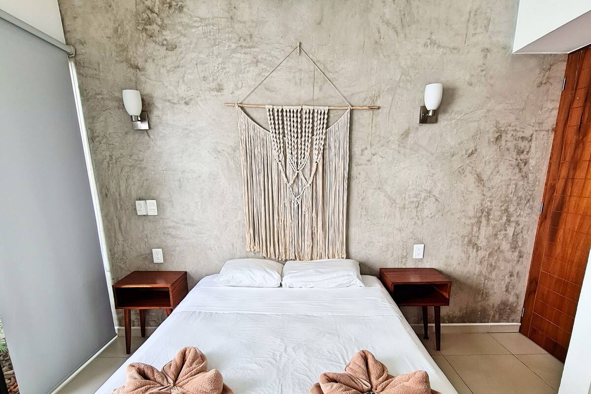 Quarto de hotel com cama, duas mesas de cabeceira, aparelho de ar condicionado e um pendente de macramé na parede.