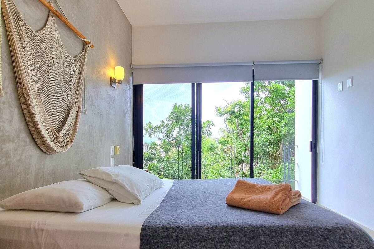 Quarto moderno com cama, rede, ventilador de teto e vista para o verde através de grandes janelas.