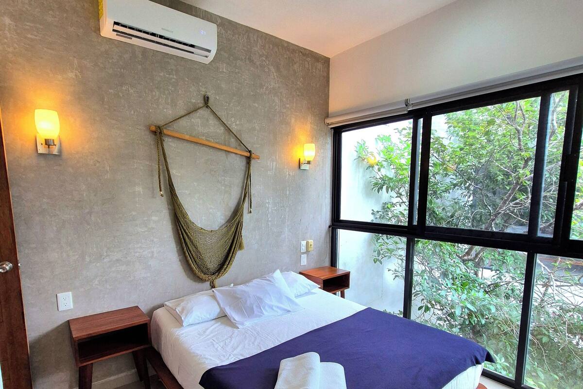 Um quarto moderno com cama, ventilador de teto, ar condicionado, rede, abajures e uma grande janela com vista para o verde.