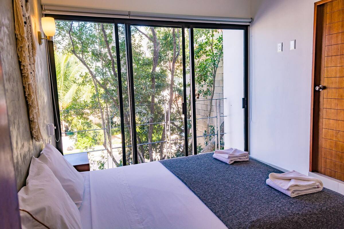 Quarto de hotel com uma cama, janela com vista para árvores e porta de madeira.