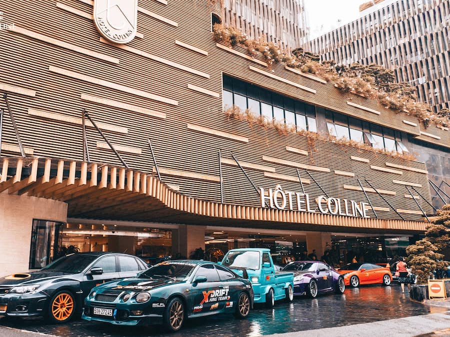 Um hotel com fachada moderna, vários carros estacionados em frente e uma placa visível com a inscrição "HOTEL COLLINE".