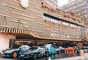 Um hotel com fachada moderna, vários carros estacionados em frente e uma placa visível com a inscrição "HOTEL COLLINE".