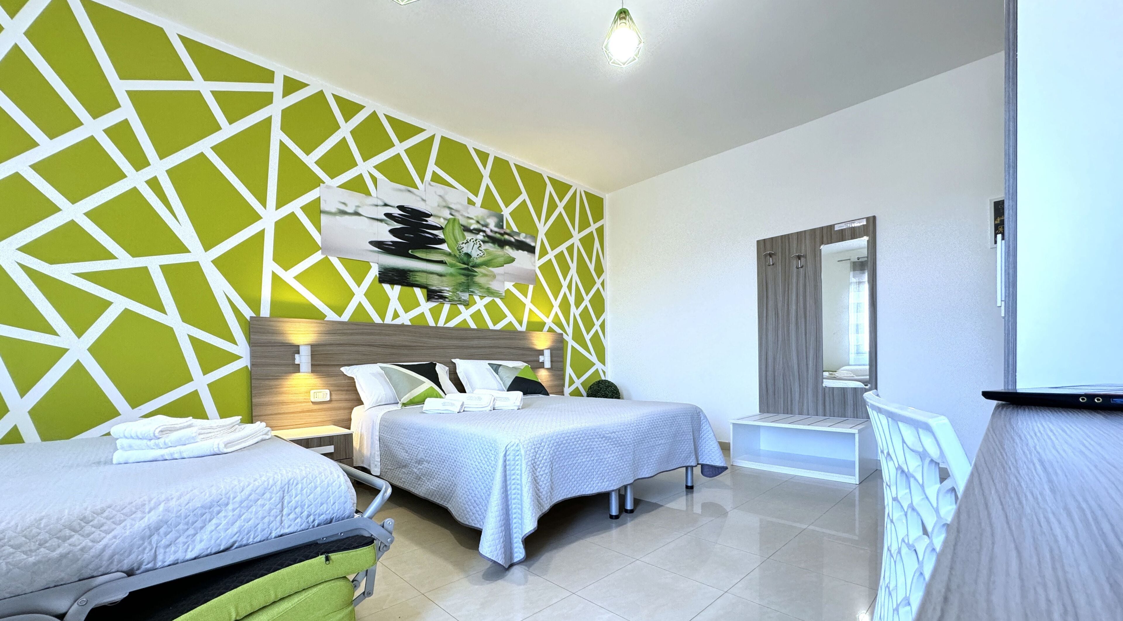 Quarto de hotel com duas camas, design geométrico verde na parede, um espelho grande e uma escrivaninha com um laptop.