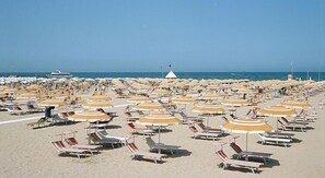 Uma praia com fileiras de espreguiçadeiras e guarda-sóis, céu claro e vista distante do mar.