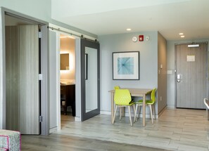 Quarto de hotel com piso de madeira, área de refeições com mesa e cadeiras, banheiro com porta-toalhas e porta que dá para outro cômodo.