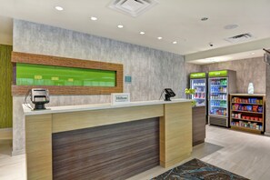 Uma área de recepção de hotel com balcão de madeira, um terminal de check-in digital, uma janela com moldura verde e máquinas de venda automática com as etiquetas 'muito gelado' e 'gelado'.