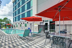 Área externa com piscina, cadeiras de sol e guarda-sóis, mesa de bilhar e um edifício moderno ao fundo.