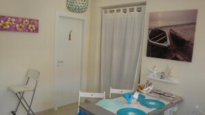 Uma área de refeições com uma mesa posta para quatro pessoas, uma cadeira dobrável branca, uma porta, uma cortina e uma parede com uma pintura de temática náutica.