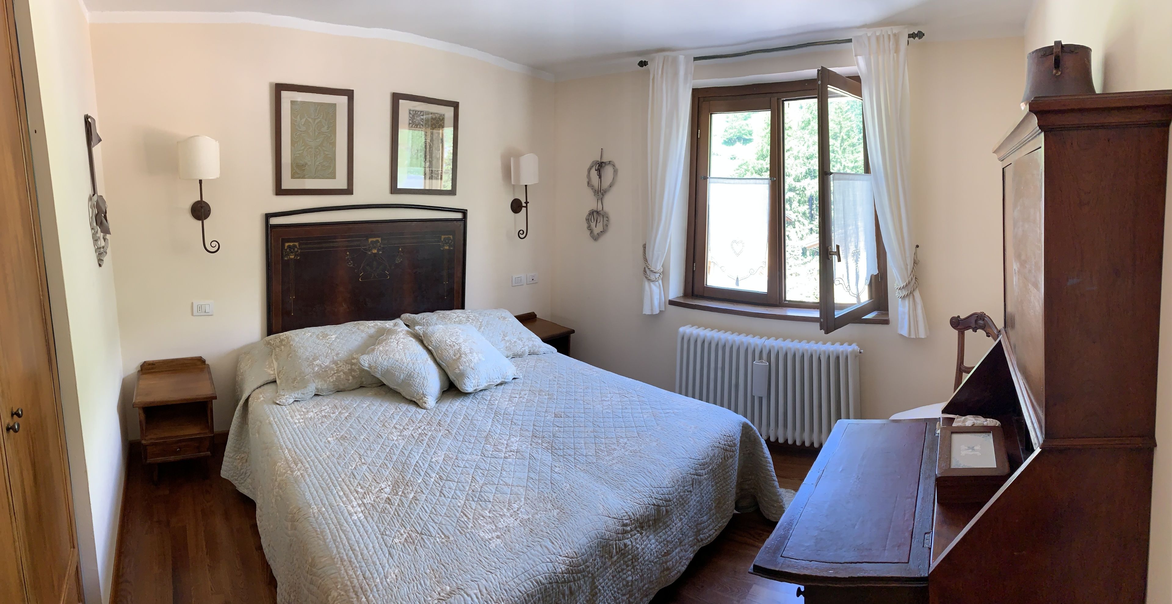 Quarto com cama, mesas de cabeceira, janela com cortinas, radiador e um armário de madeira.