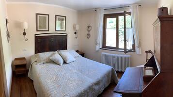 Quarto com cama, mesas de cabeceira, janela com cortinas, radiador e um armário de madeira.