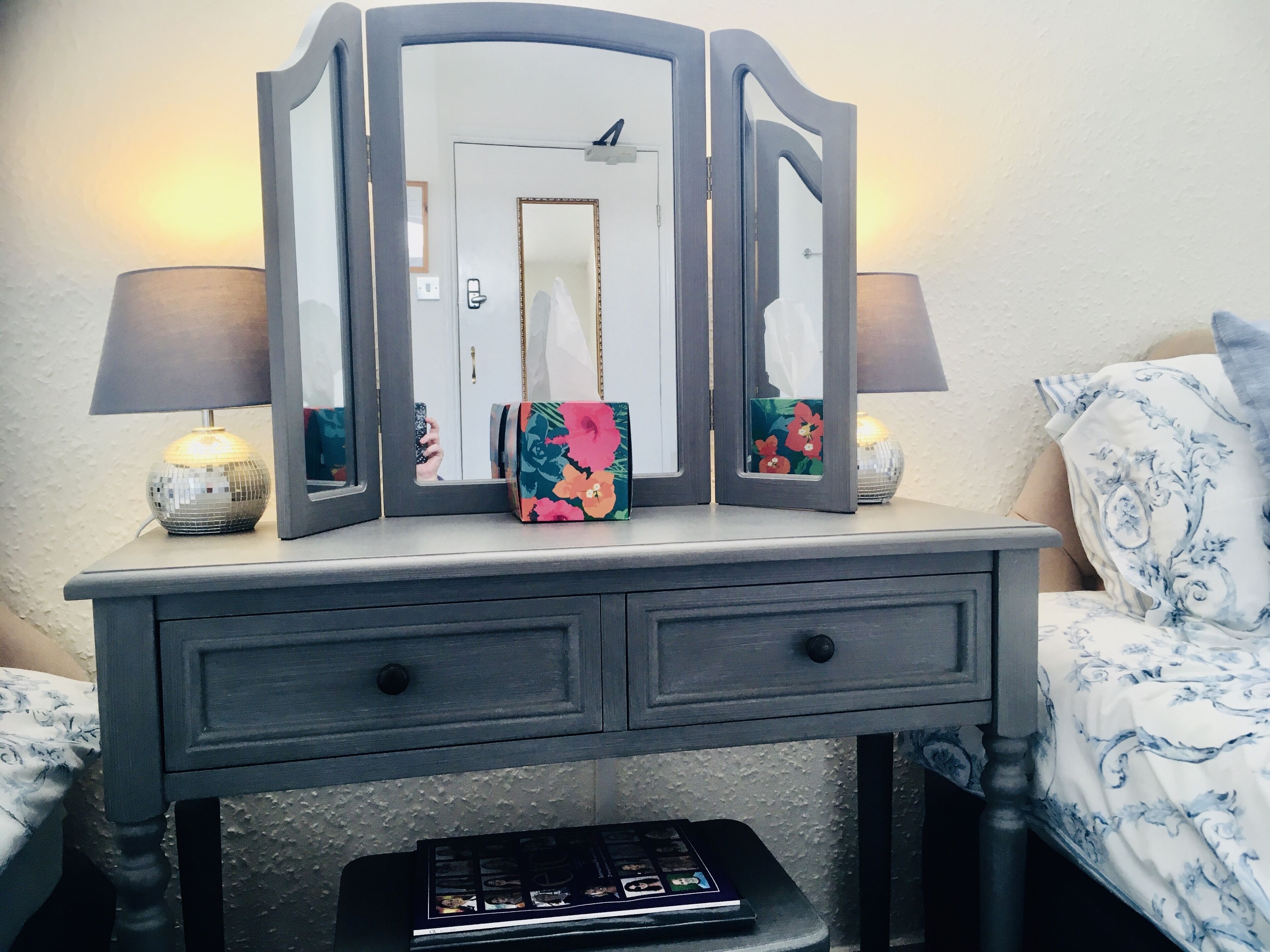 Uma toucador com espelho, duas luminárias e uma pequena mesa com um objeto decorativo.