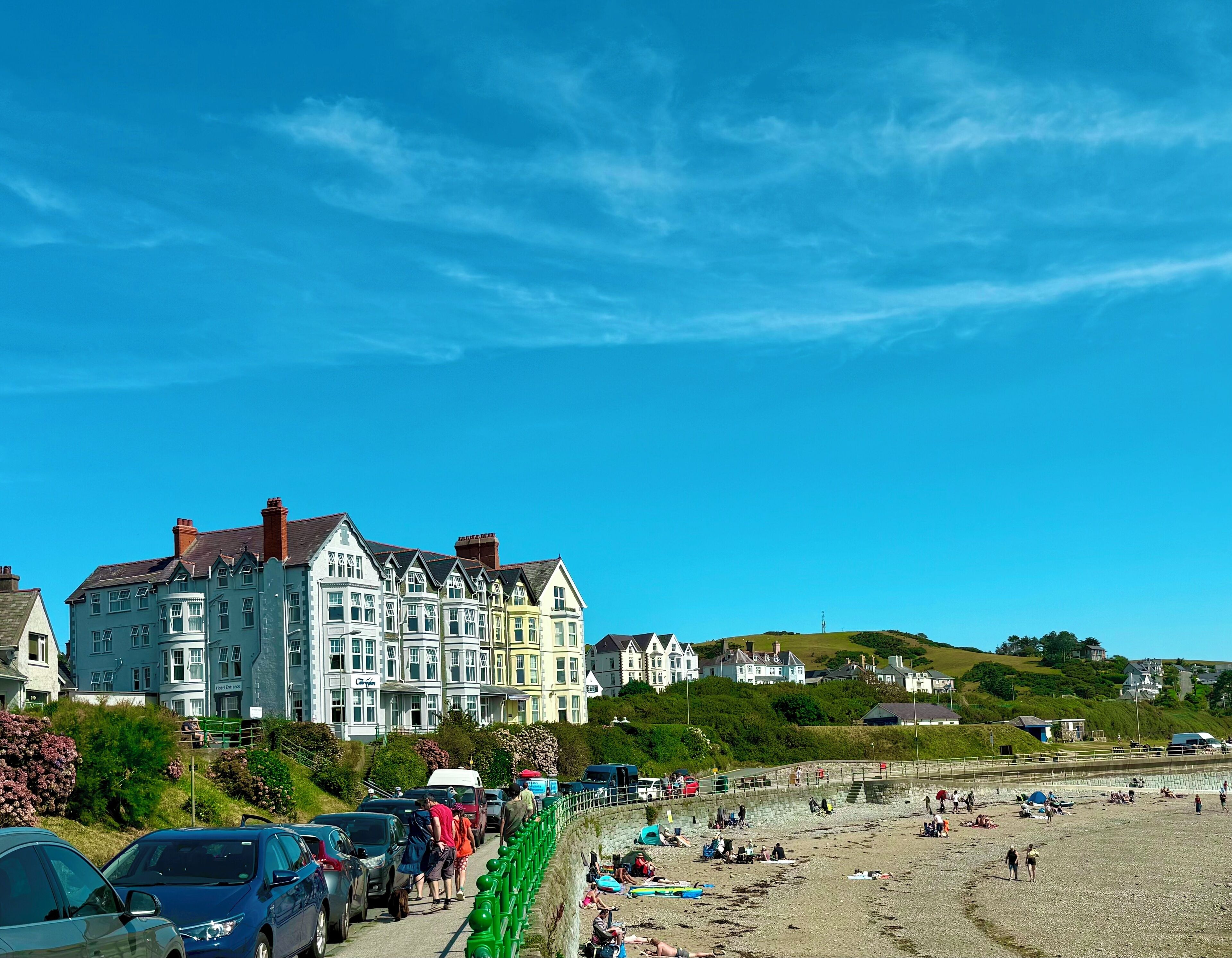 Uma cidade costeira com uma fileira de edifícios coloridos em estilo vitoriano, uma praia de areia com pessoas e carros estacionados ao longo do calçadão.