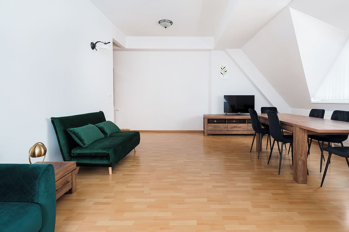 Quarto moderno e bem iluminado com uma mesa de jantar de madeira, cadeiras pretas, sofá verde e uma televisão em um armário de madeira.