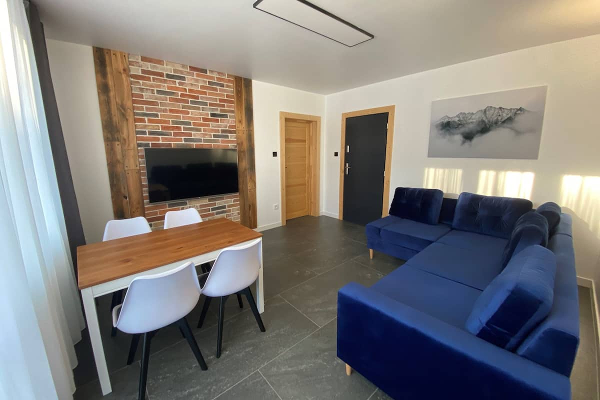 Uma sala de estar moderna com uma parede de destaque em tijolo, uma televisão de tela plana grande, um sofá modular azul e uma mesa de jantar de madeira com cadeiras brancas.