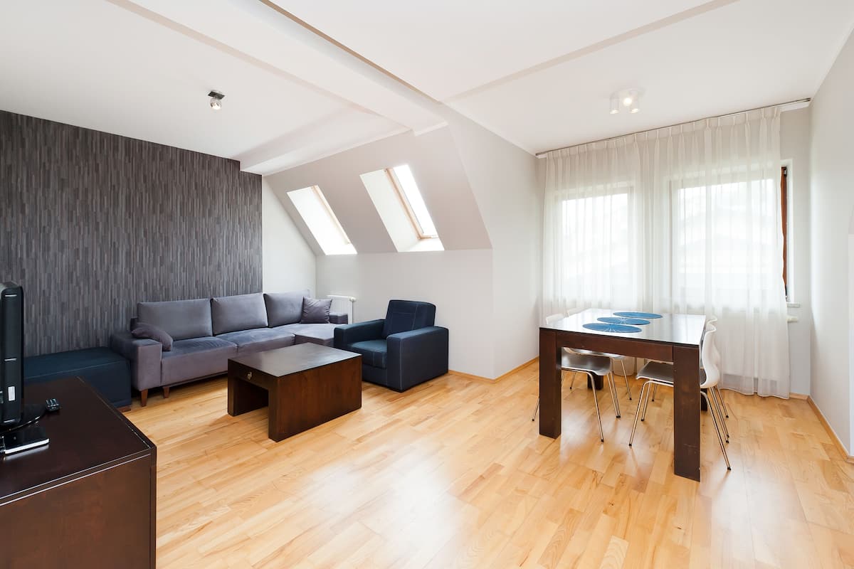 Uma sala de estar moderna com sofá, mesa de centro e mesa de jantar com cadeiras. Há claraboias e uma grande janela com cortinas.