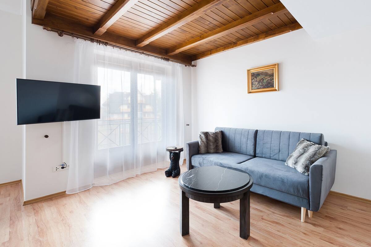 Uma sala de estar com sofá azul, mesa de centro preta, TV fixada na parede e uma janela com cortinas leves.