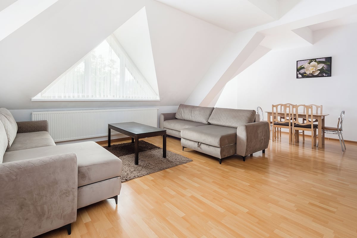 Apartamento amplo no estilo loft, com pé-direito alto, uma grande janela, um conjunto de sofás, uma mesa de jantar com cadeiras e um radiador.
