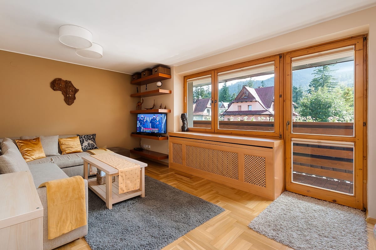 Uma sala de estar com sofá, mesa de centro, televisão e uma janela grande com vista para casas.