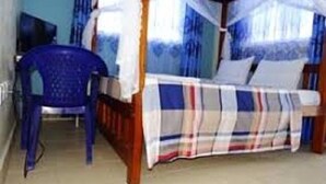 Uma cama de solteiro com colcha xadrez, cabeceira de madeira e dossel. Há uma cadeira de plástico azul e uma televisão no quarto.