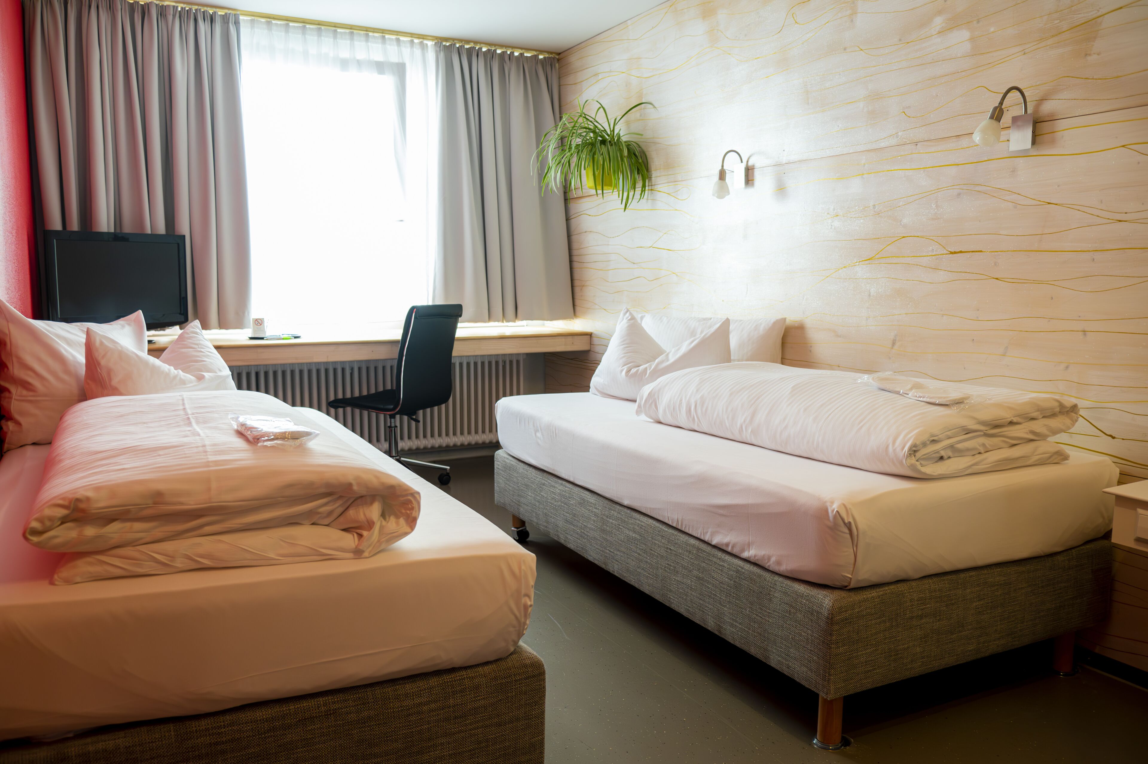 Quarto de hotel com duas camas, uma escrivaninha com computador, uma cadeira, um radiador e uma planta.