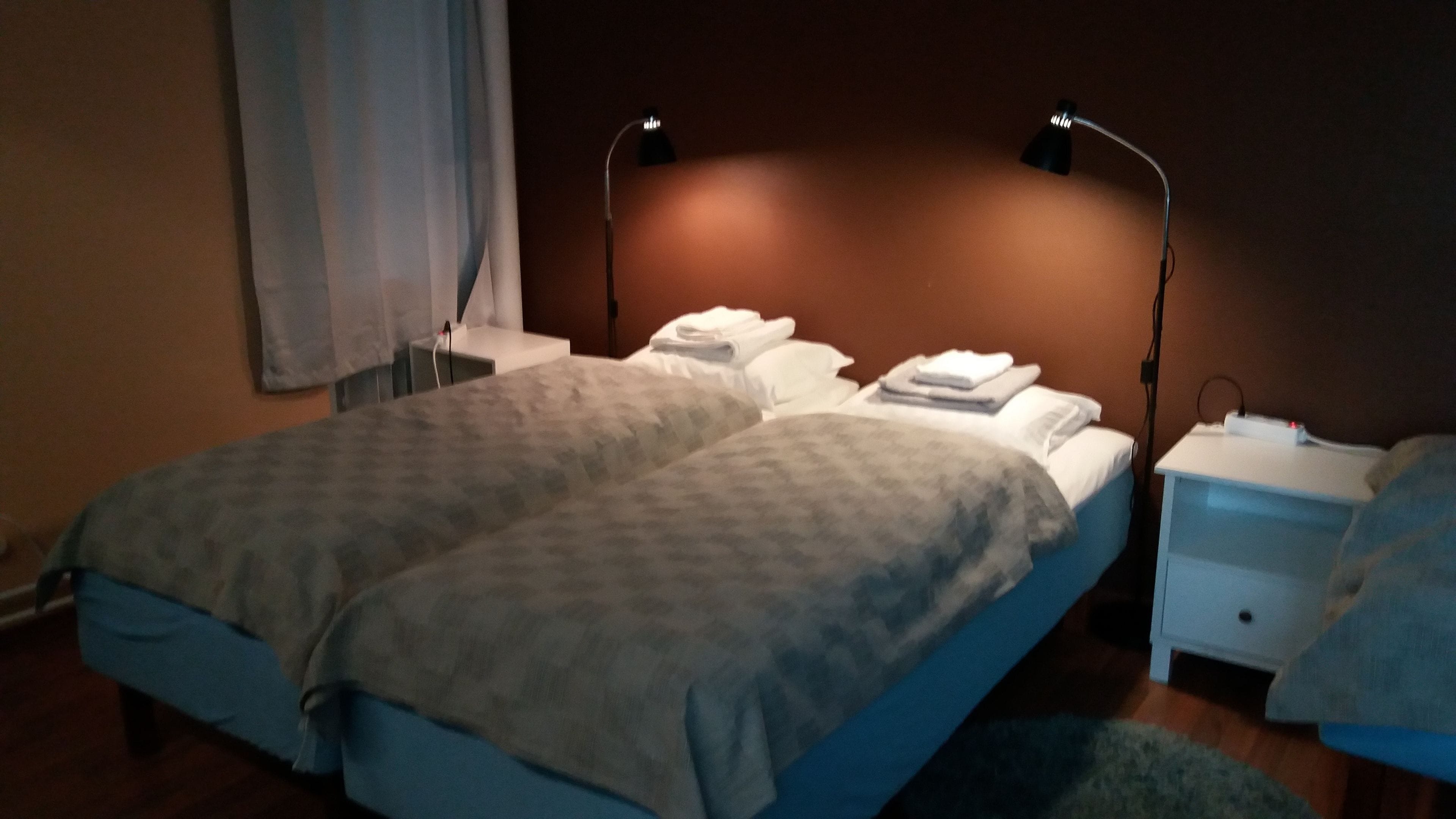 Uma cama de casal com edredom cinza, duas mesas de cabeceira, um criado-mudo e uma luminária, com uma régua de tomadas no lado direito.
