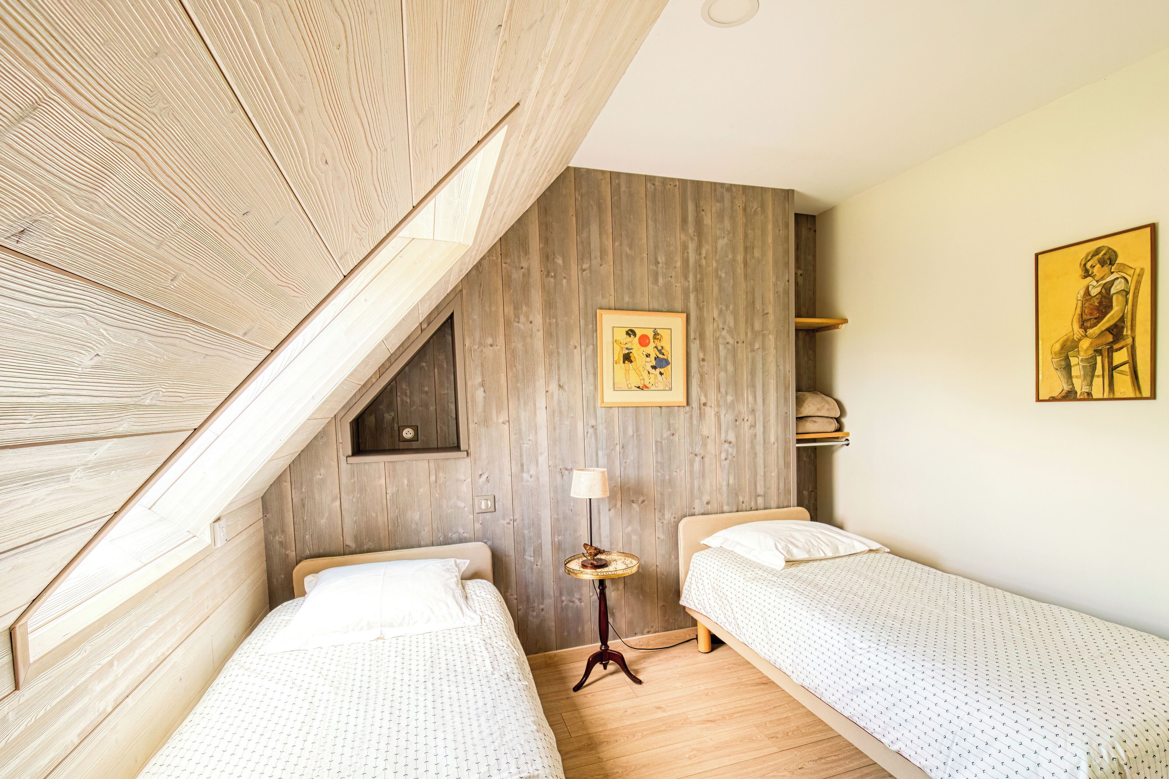 Quarto com duas camas de solteiro, paredes de madeira, uma mesa redonda pequena com abajur e um quadro na parede.