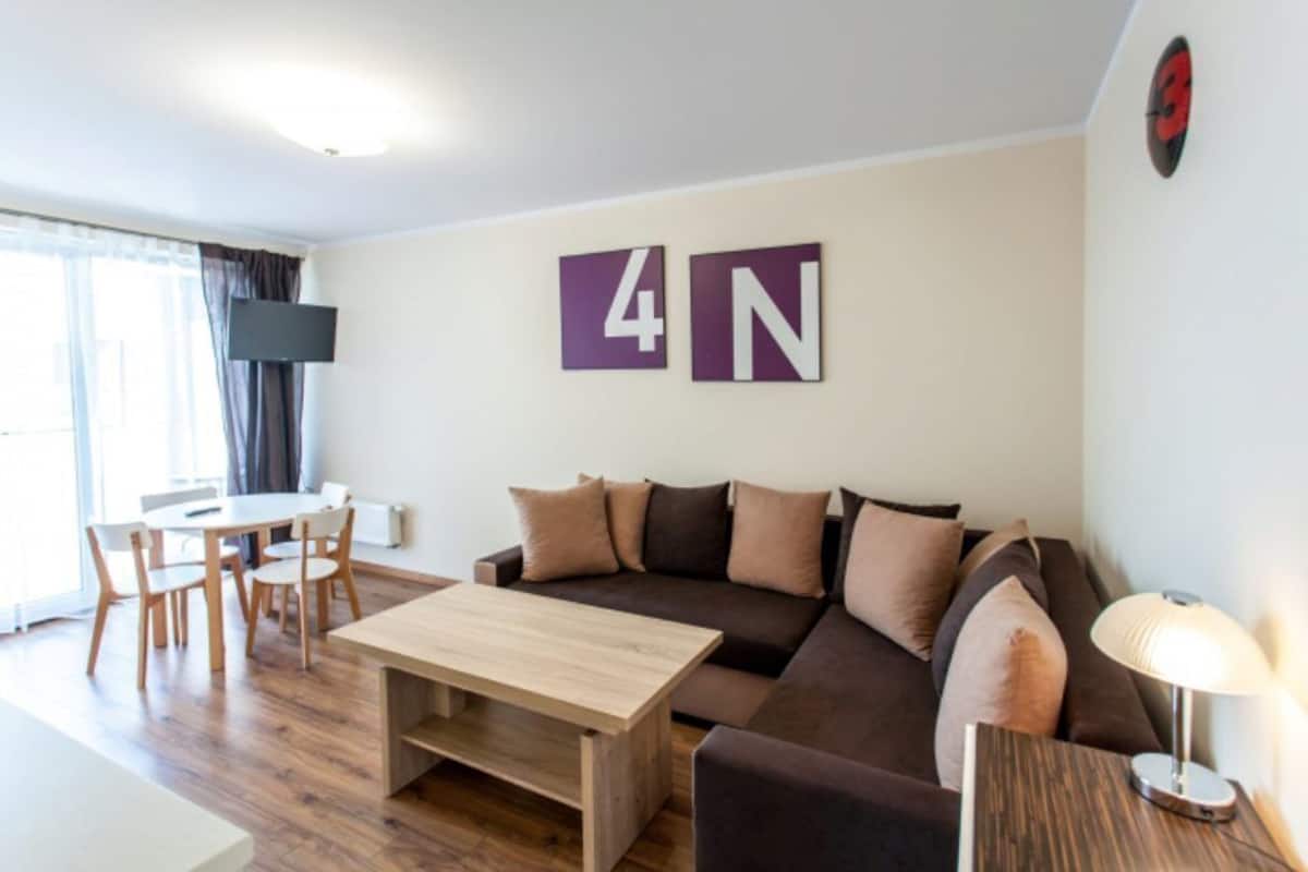 Uma sala de estar moderna com sofá, mesa de centro e um espaço de jantar com cadeiras e mesa. Há uma TV na parede e uma placa com as letras "4N".