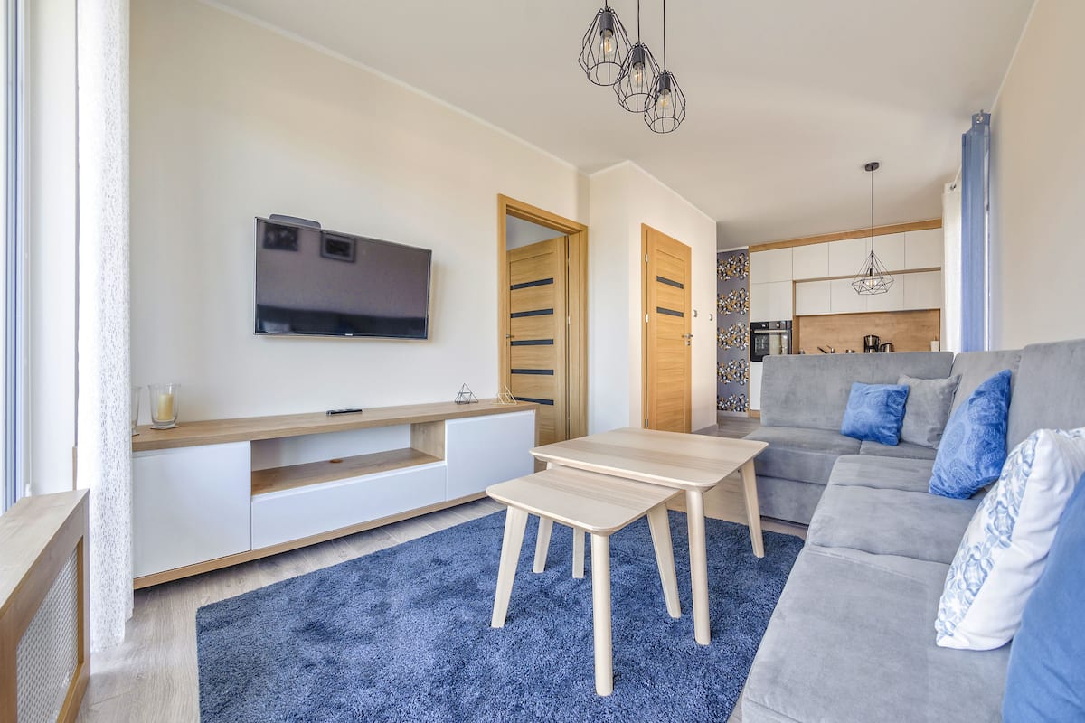Uma sala de estar moderna com um sofá cinzento, mesa de centro de madeira e uma TV de tela plana fixada na parede.